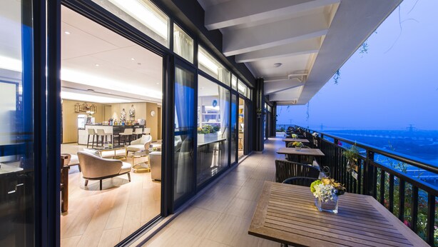 Varanda de hotel moderna com vista para o mar, mobiliada com mesas e cadeiras de madeira, e uma área de refeições visível através de grandes portas de vidro.