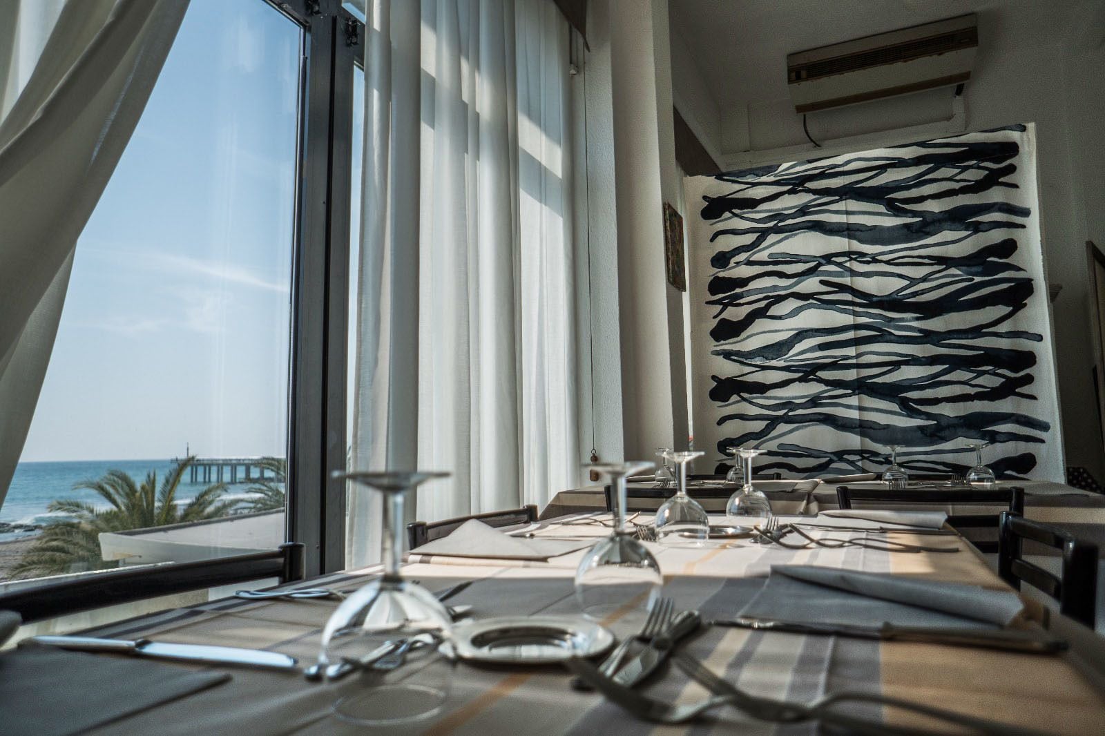 Mesa de jantar posta com talheres e copos de vinho, com vista para uma praia e um cais.