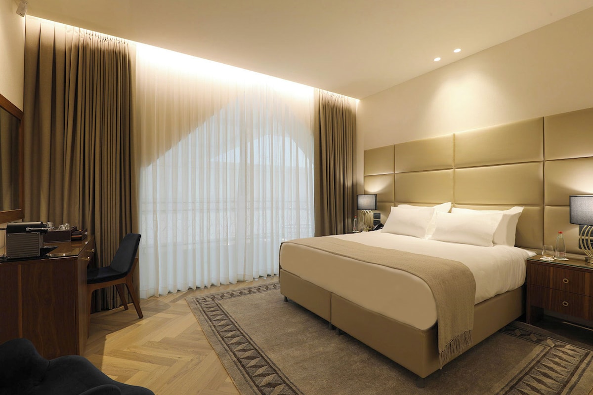 Quarto de hotel com uma cama grande, uma escrivaninha, uma cadeira e uma mesinha de cabeceira.