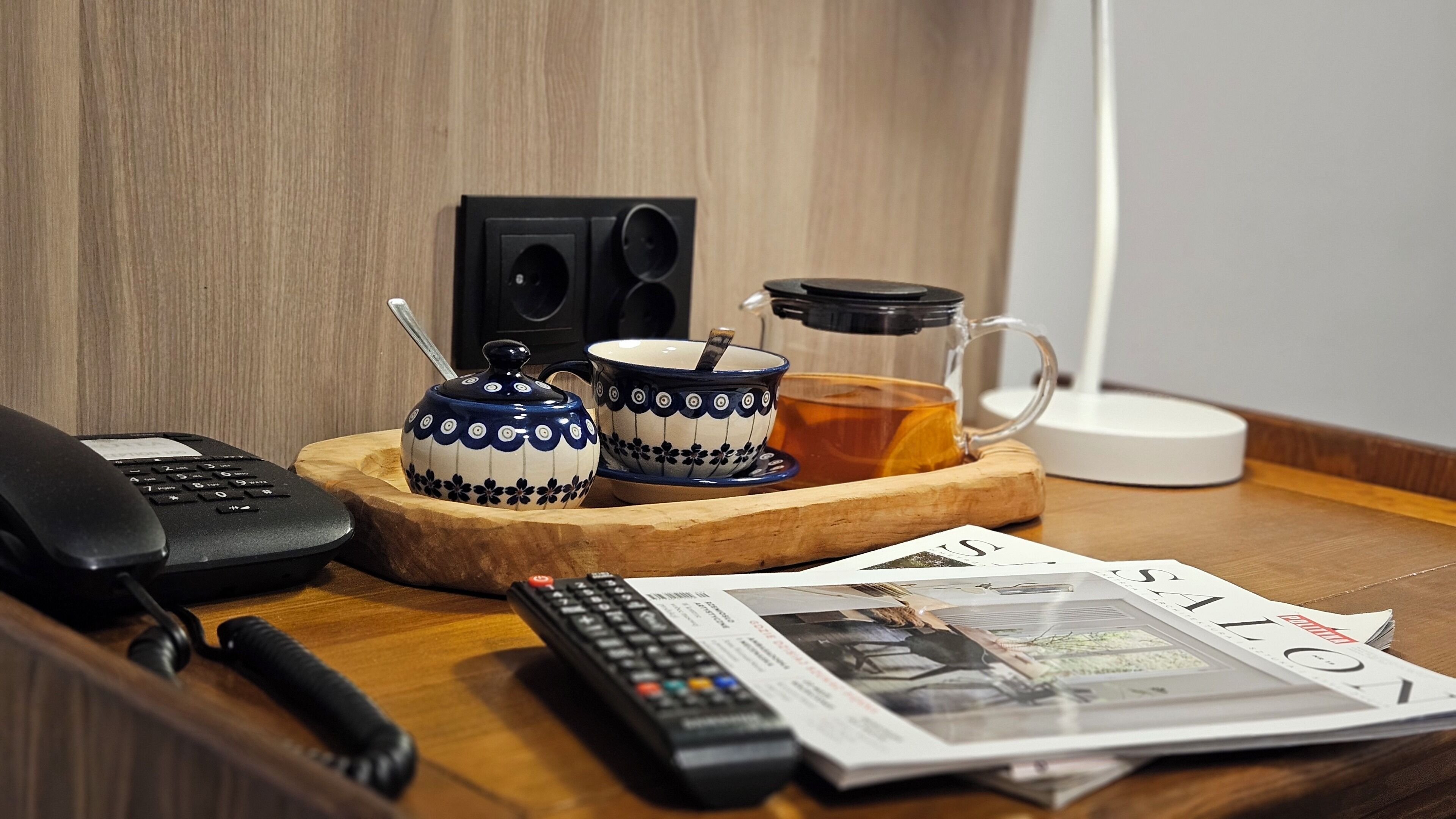Uma mesa de madeira com telefone, duas xícaras, uma cafeteira, um controle remoto e um jornal.