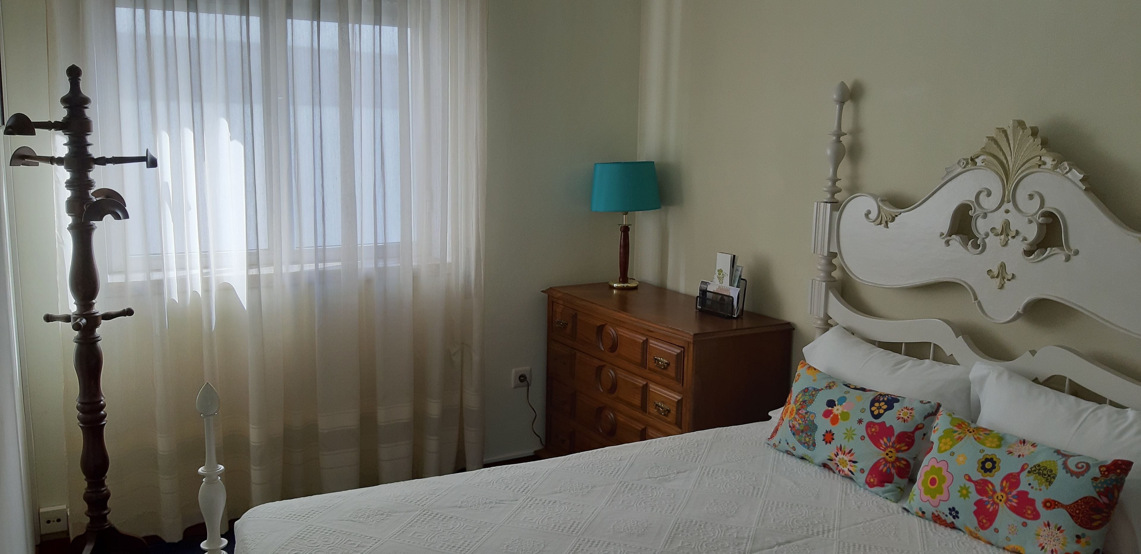 Quarto com cama, penteadeira, luminária e janela com cortinas leves.