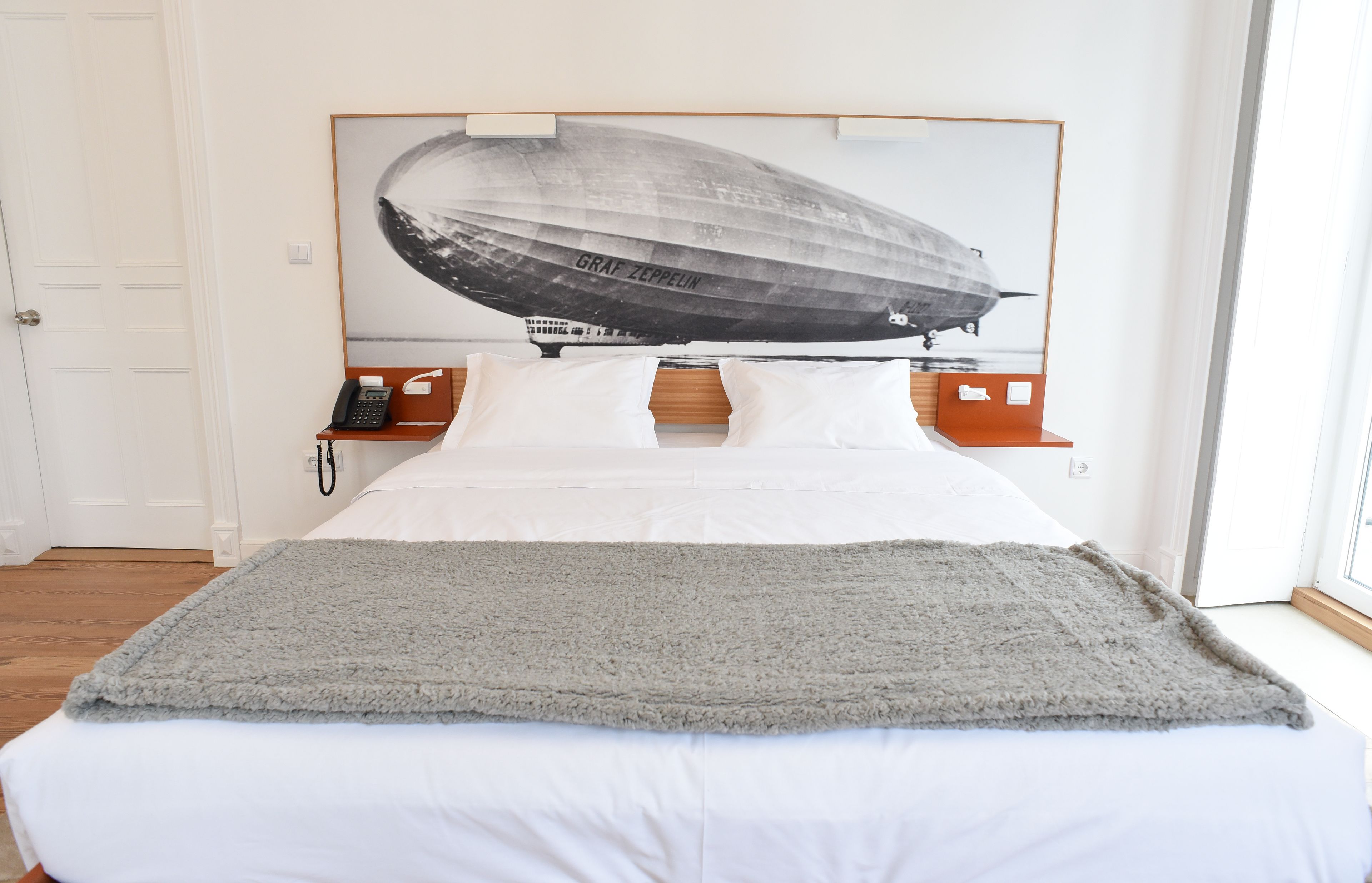 Uma cama bem arrumada com um cobertor cinzento, um travesseiro branco e uma grande obra de arte com o tema Zeppelin na cabeceira.