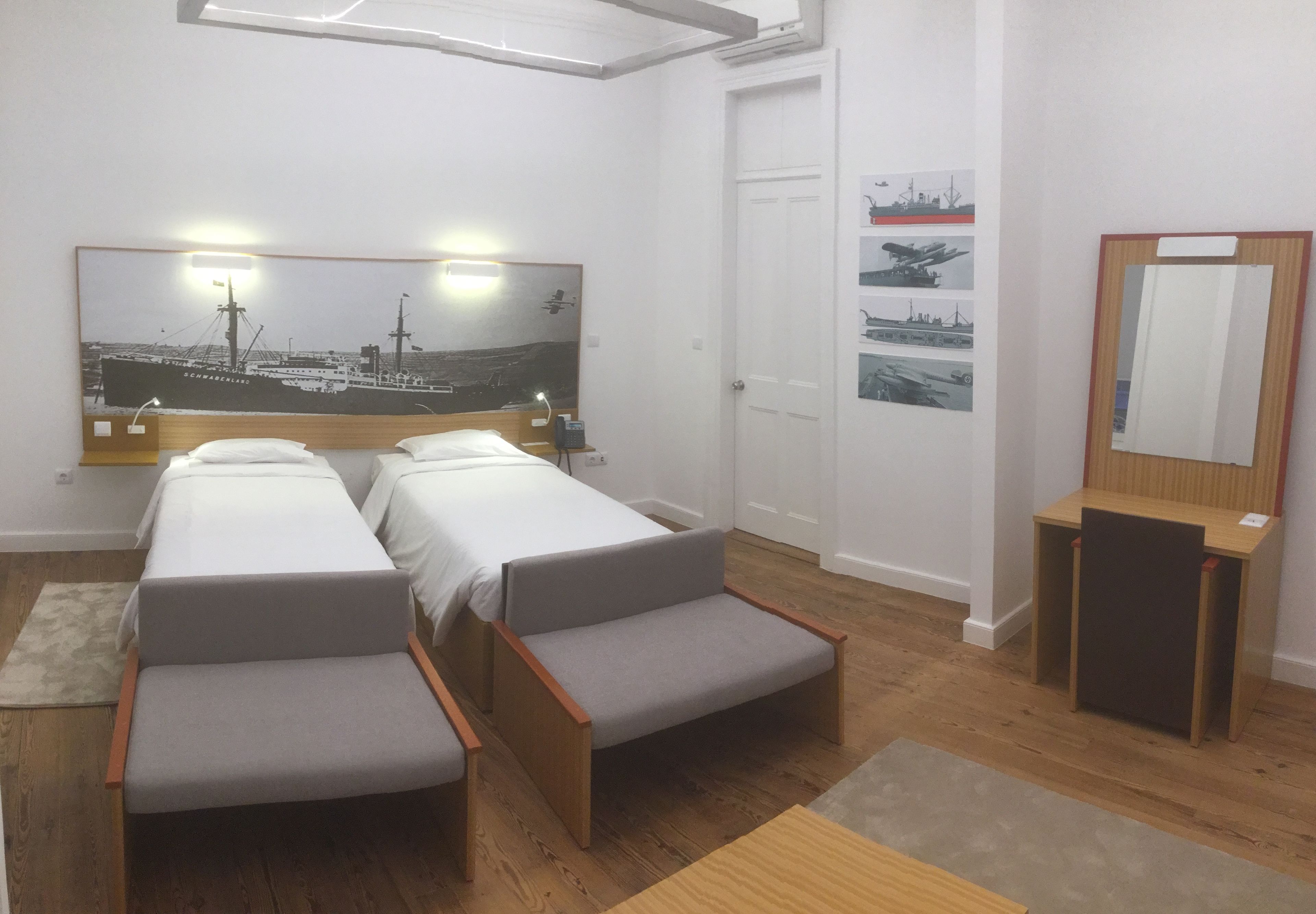 Quarto com duas camas de solteiro, uma escrivaninha de madeira, um espelho e uma grande moldura com a imagem de um navio.