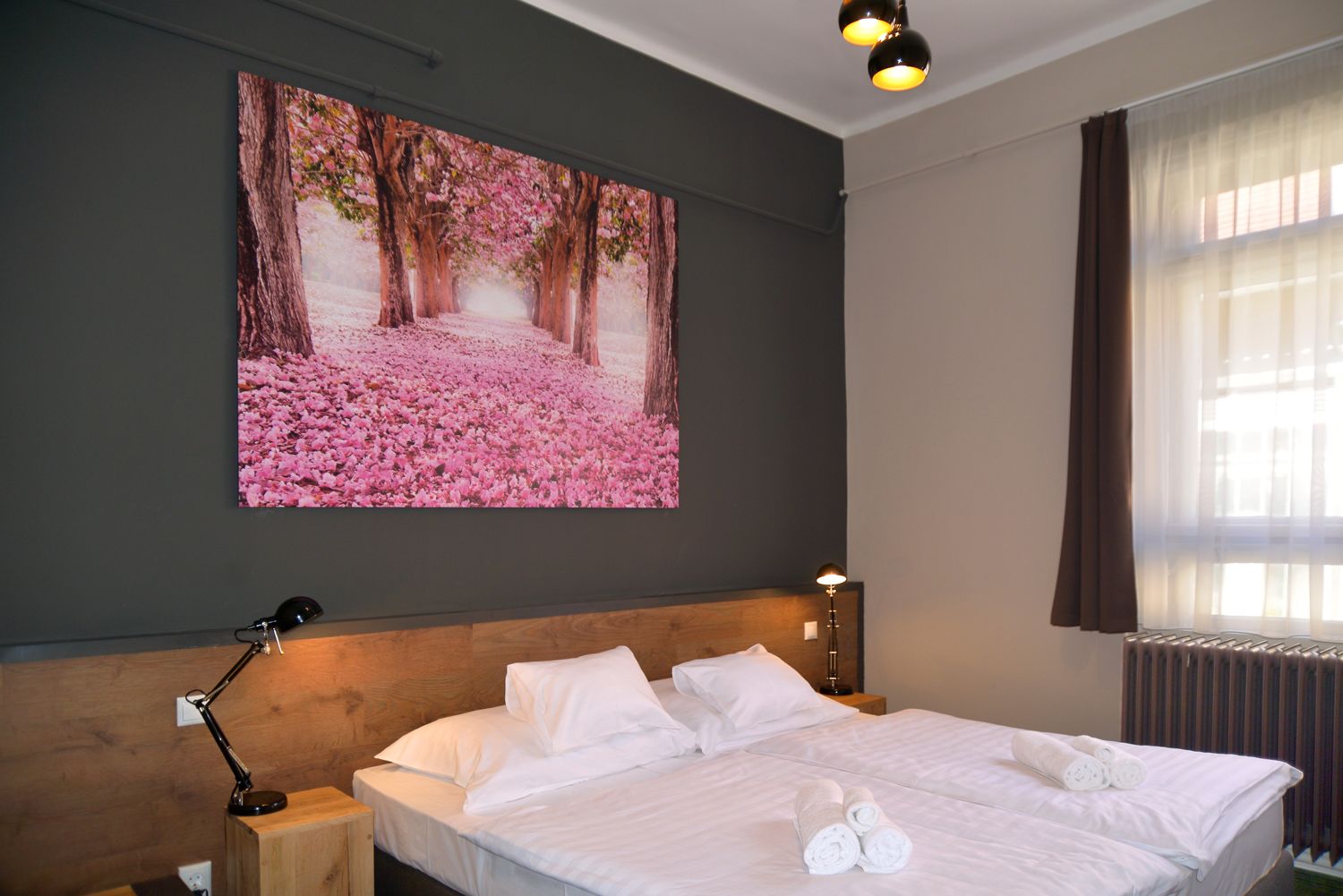 Quarto de hotel com uma cama grande, mesa de cabeceira, luminária de mesa, luminária de parede, radiador e uma janela ampla com cortinas.