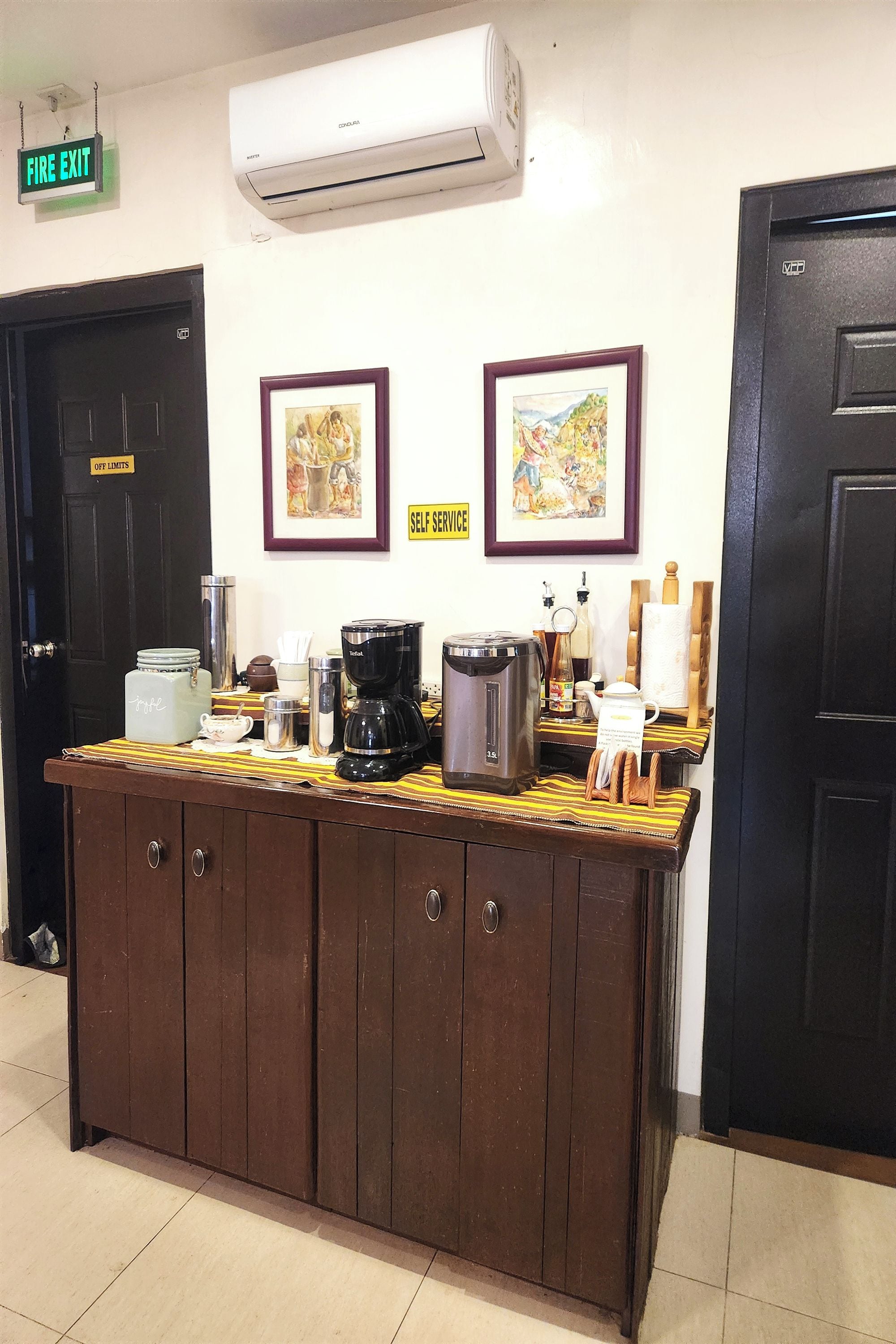 Uma cozinha compacta com cafeteira, chaleira e chá. Há duas quadros na parede, uma placa de saída de incêndio e uma placa de autosserviço.