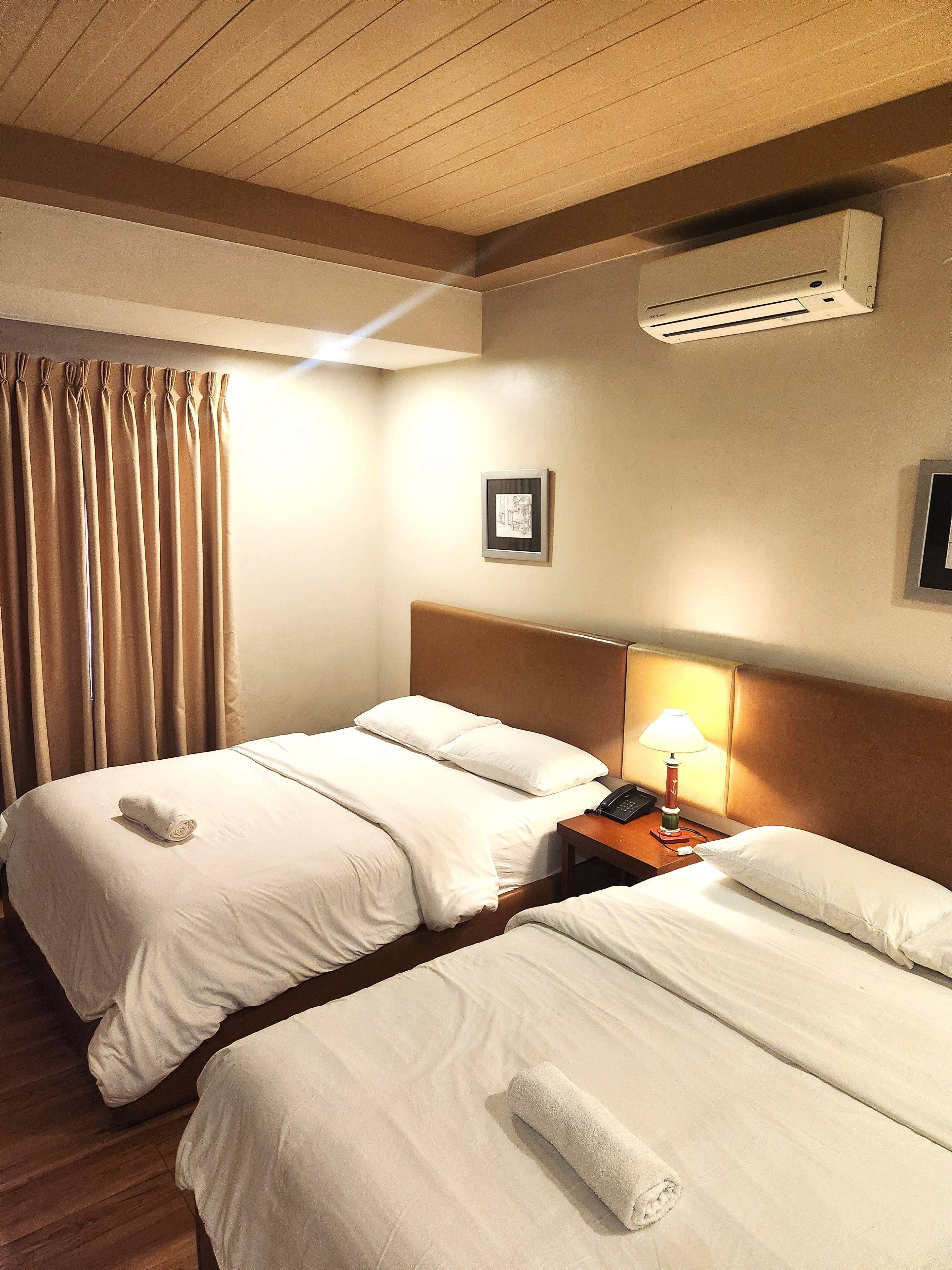 Quarto de hotel com duas camas, mesa de cabeceira com abajur, ar condicionado e um quadro na parede.