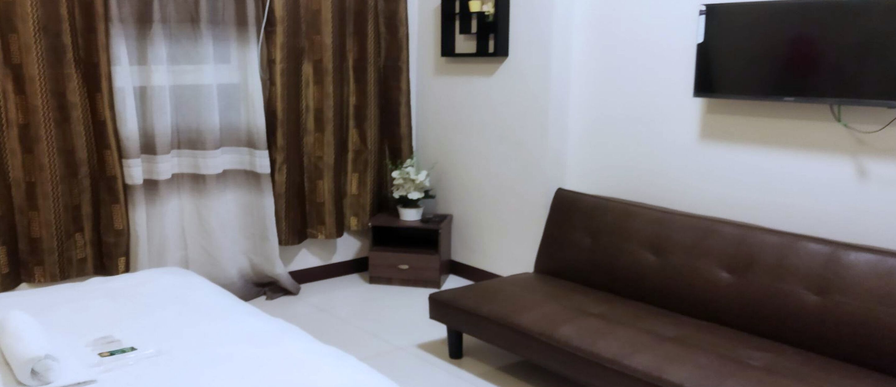 Quarto de hotel com cama, um sofá marrom, uma mesinha com planta em vaso, televisão na parede e janela com cortinas.