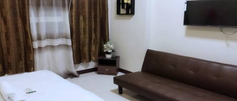 Quarto de hotel com cama, um sofá marrom, uma mesinha com planta em vaso, televisão na parede e janela com cortinas.