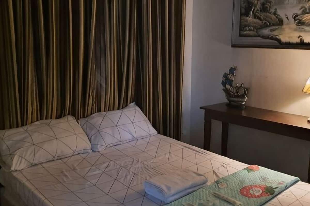Uma cama com edredom estampado em padrão geométrico, uma toalha dobrada e uma almofada decorativa. Uma mesa com abajur e uma tigela são visíveis ao fundo.