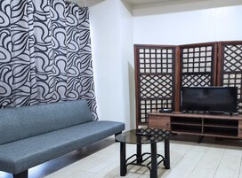 Uma sala de estar com um sofá cinzento, uma mesa de centro de vidro, uma televisão sobre um suporte de madeira e um biombô decorativo com padrão geométrico.
