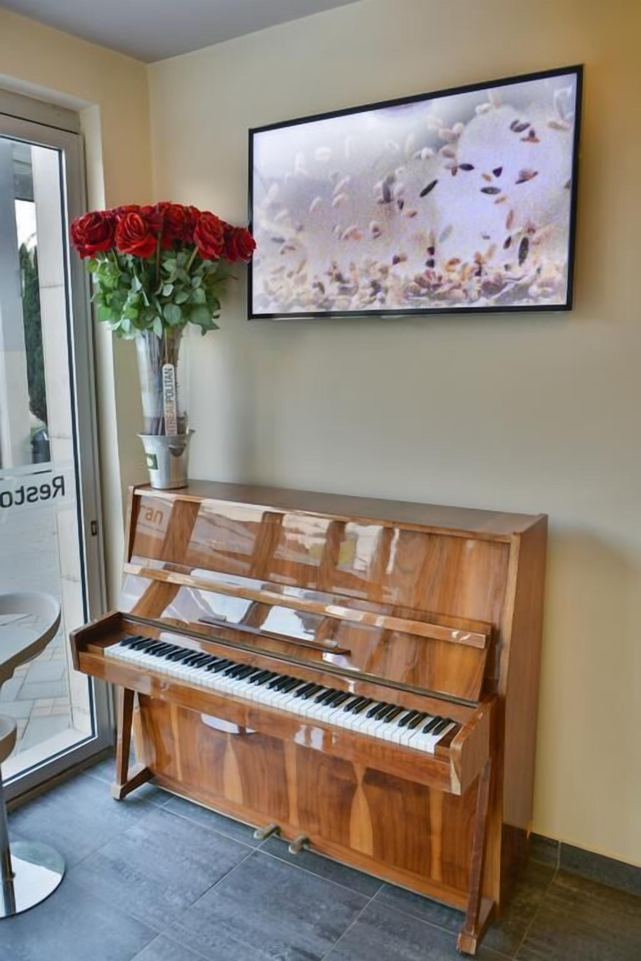 Um piano de madeira num quarto com uma janela grande, um vaso de rosas vermelhas e um quadro na parede.