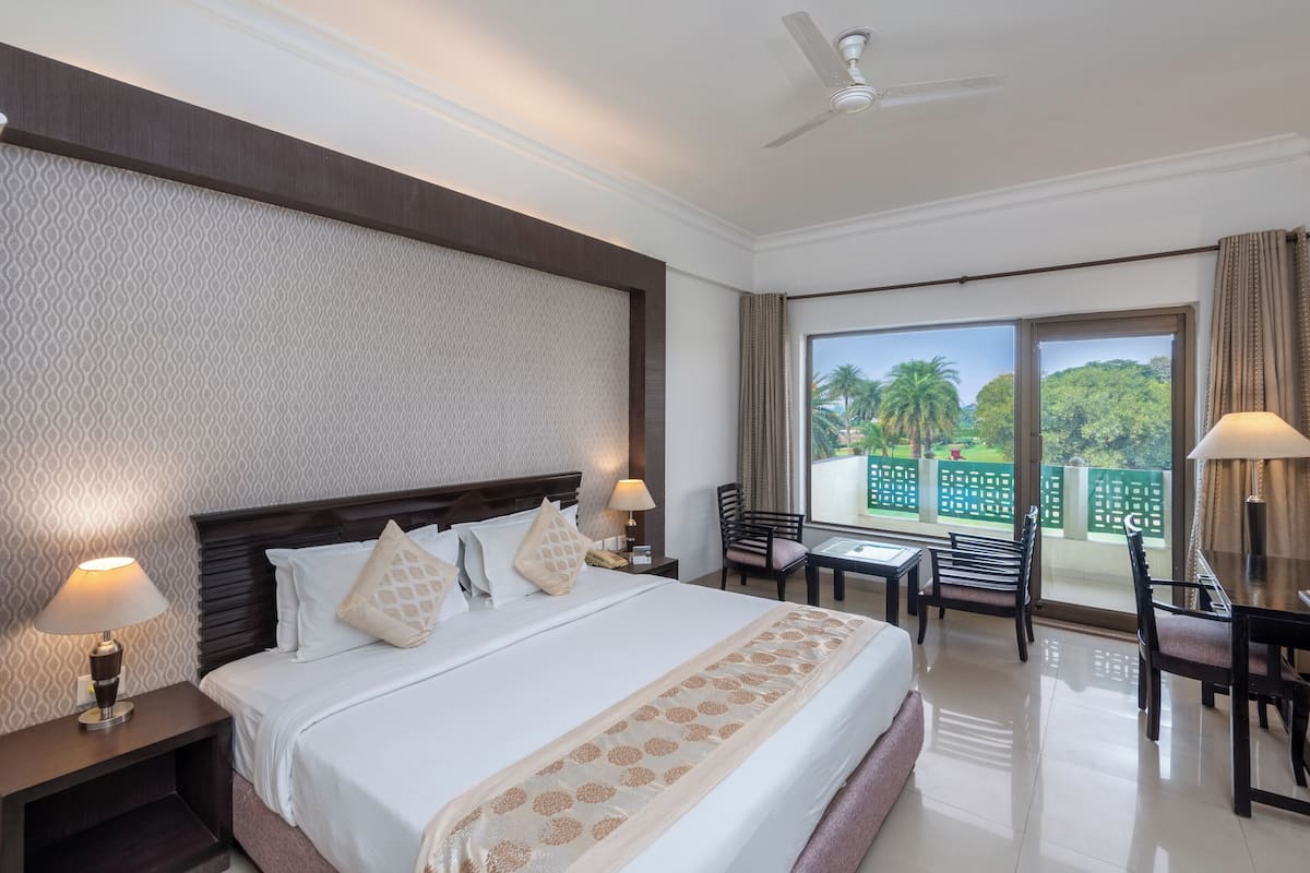 Quarto de hotel com cama grande, duas mesas de cabeceira com luminárias, ventoinha no teto, varanda com vista para palmeiras e área de refeições com cadeiras e mesa.