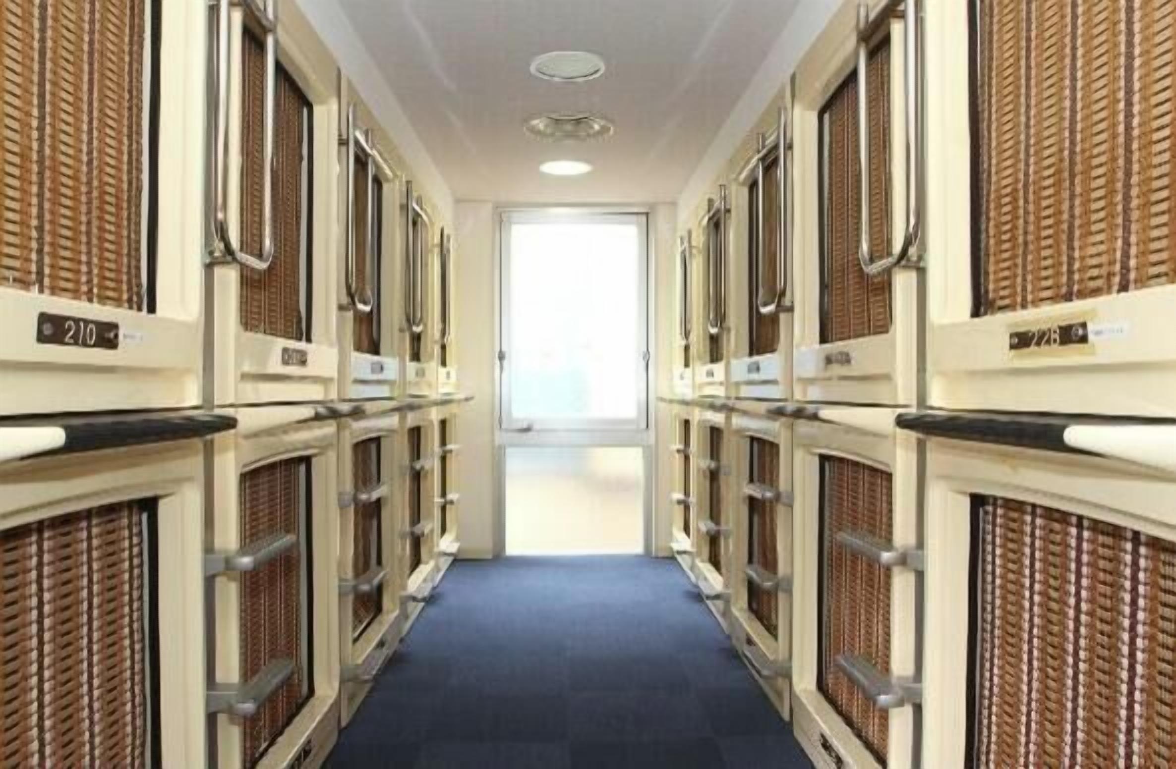 Um corredor com fileiras de quartos, cada um com uma placa de número, que leva a uma saída iluminada.