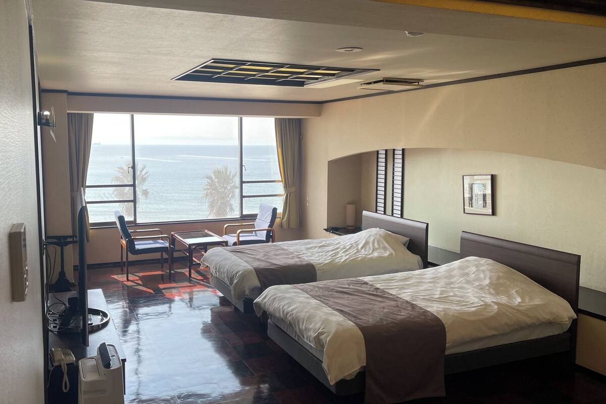 Quarto de hotel com duas camas, uma mesa, uma cadeira e uma janela grande com vista para o mar.