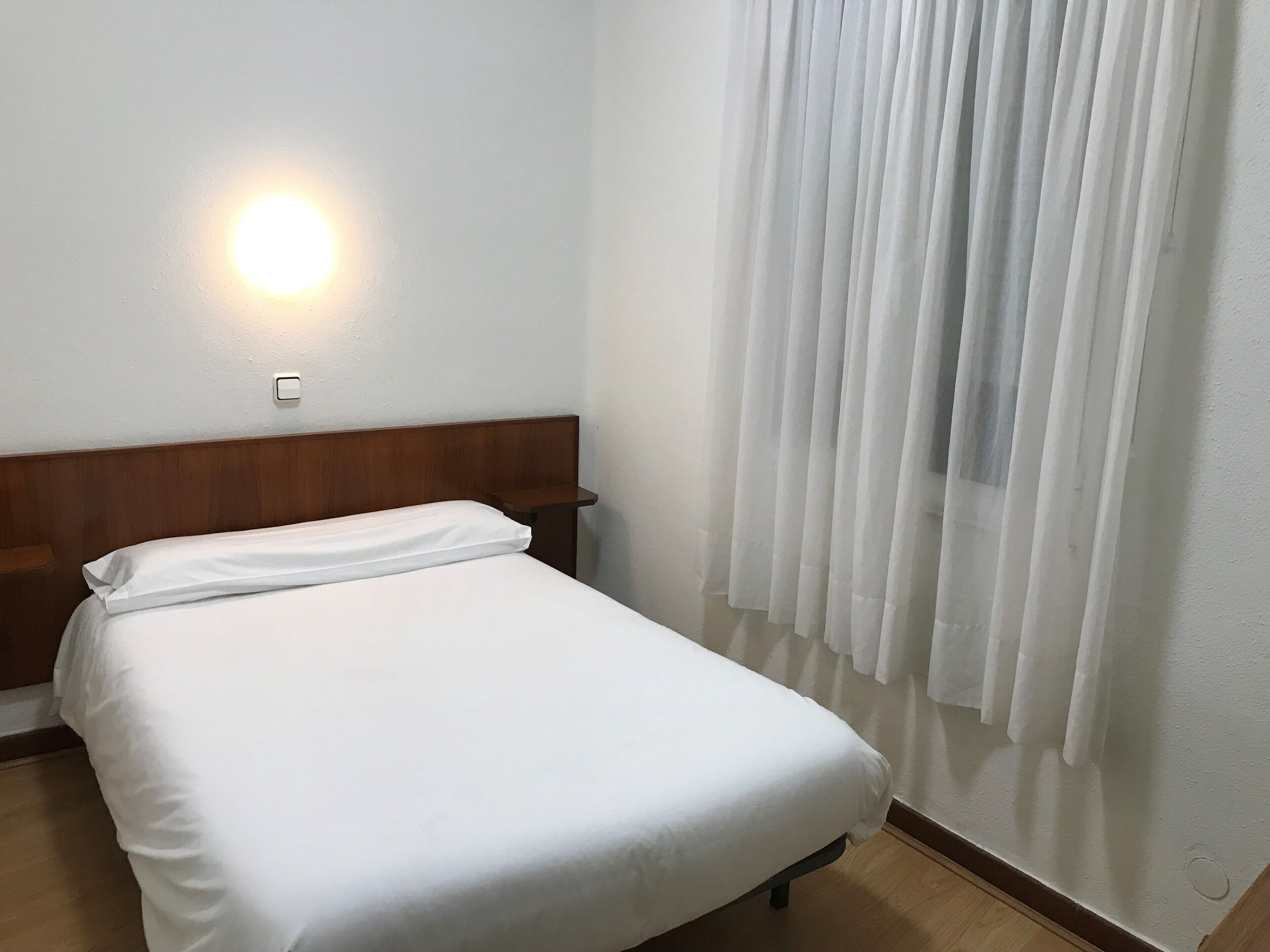 Uma cama bem arrumada num quarto de hotel, com cabeceira de madeira, mesa de cabeceira e luminária na parede.