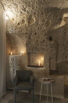 Quarto com parede de pedra texturizada, uma poltrona acolchoada, uma pequena mesa com uma revista e duas velas acesas em nichos embutidos.