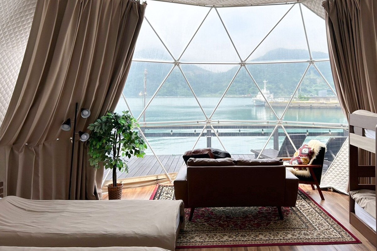 Uma cabana acolhedora com uma grande janela que oferece vista para um corpo de água e uma ponte. No interior, há duas camas, um sofá e uma mesinha com uma planta.