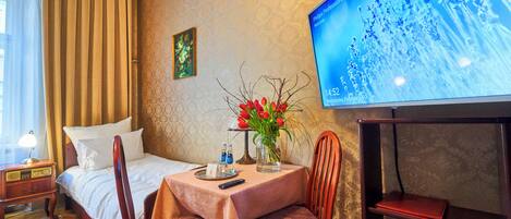Quarto de hotel com cama, escrivaninha com luminária, televisão, mesa com um vaso de flores e uma cadeira.