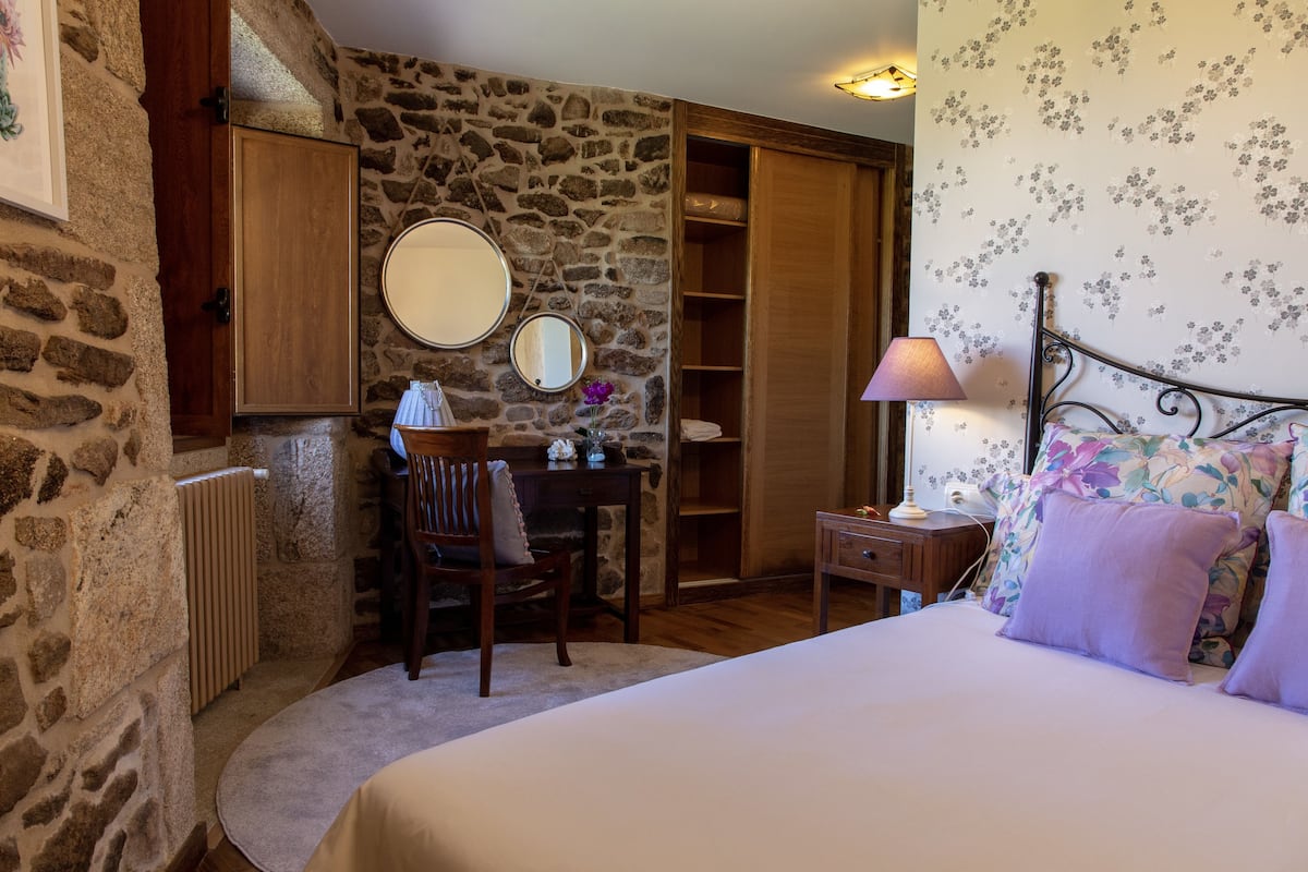 Quarto com parede de pedra, móveis de madeira, cama com roupa de cama florida e mesa de cabeceira com abajur.