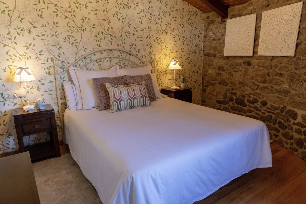Quarto com parede de pedra, cama com roupa de cama branca, duas mesas de cabeceira com abajures e papel de parede floral.