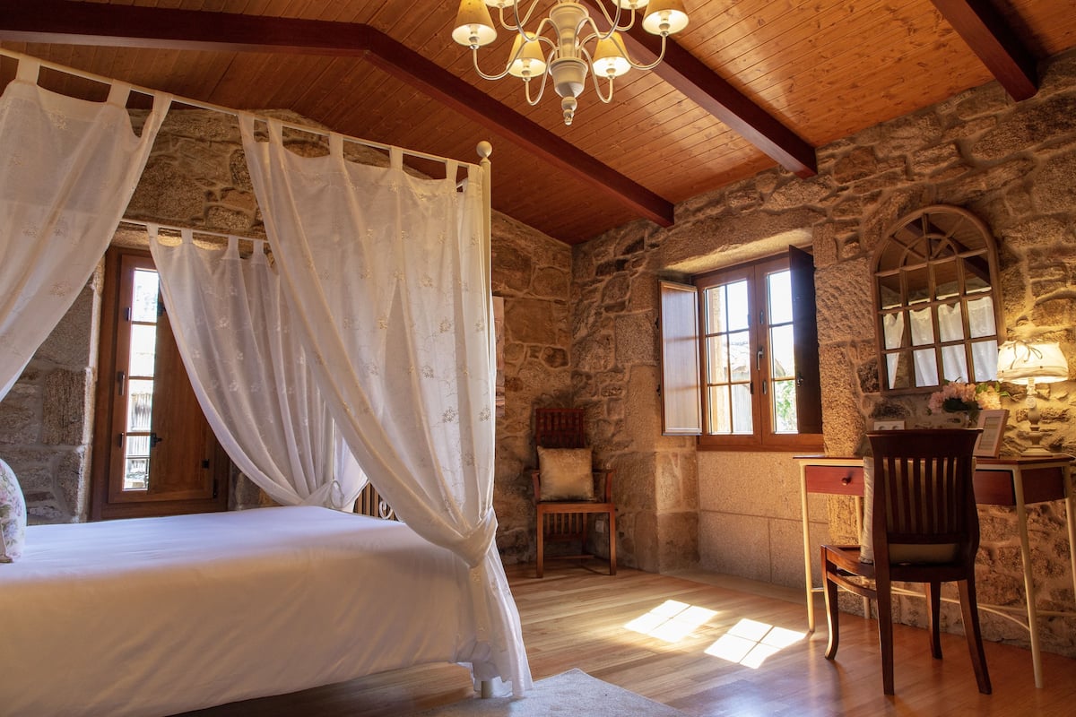 Quarto com cama de dossel, parede de pedra, vigas de madeira, escrivaninha com cadeira e janela com vista.