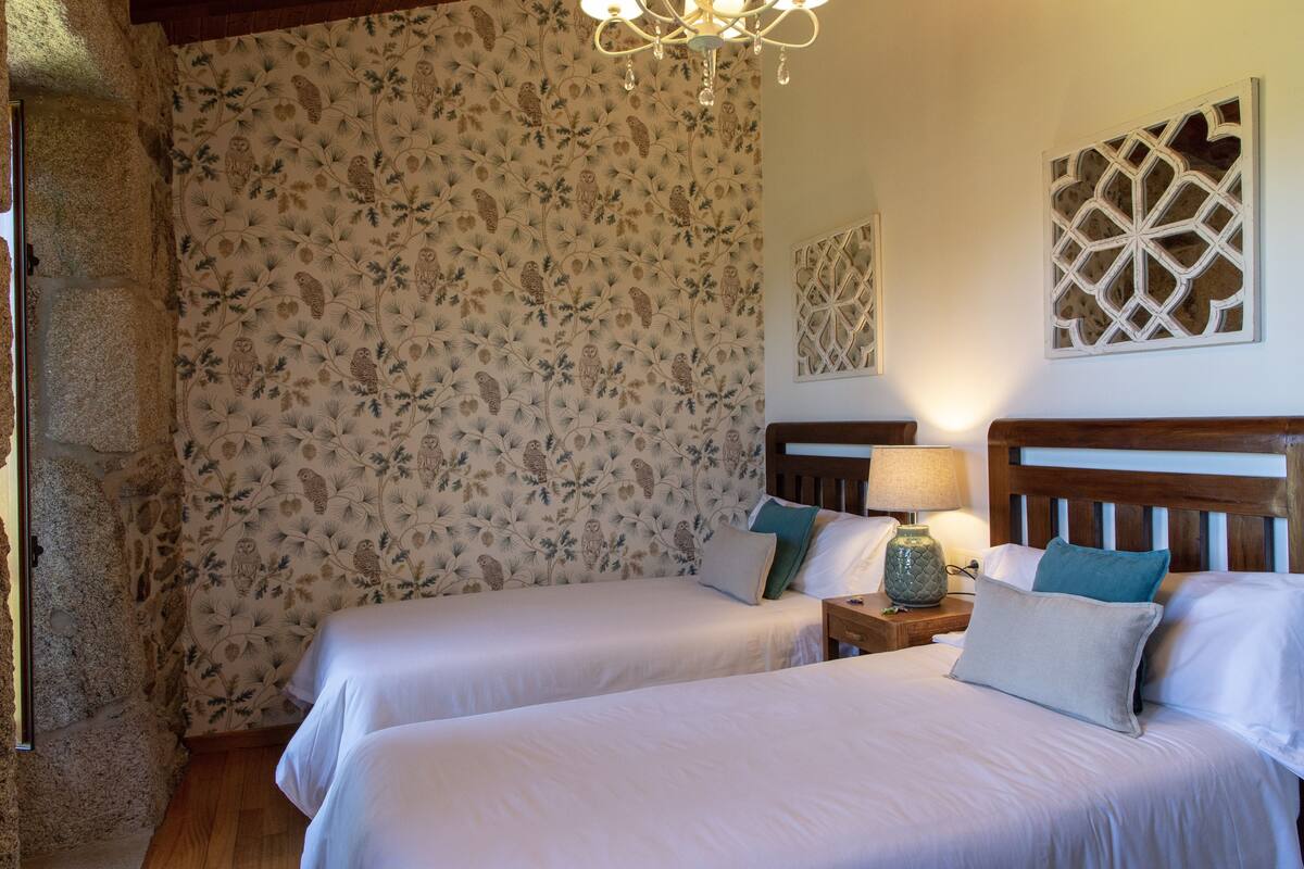 Quarto com duas camas, cabeceira de madeira, mesa de cabeceira com abajur e um painel decorativo na parede.
