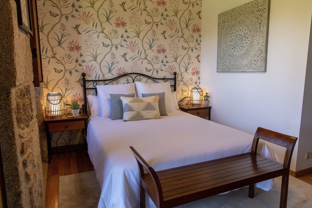 Uma cama bem arrumada com um banco de madeira, uma mesa de cabeceira com abajur e um papel de parede floral.