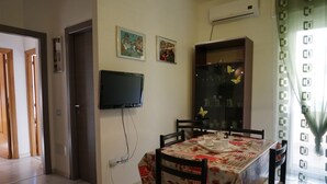 Uma área de refeições com uma mesa posta para quatro pessoas, uma televisão fixada na parede e uma estante com objetos decorativos.