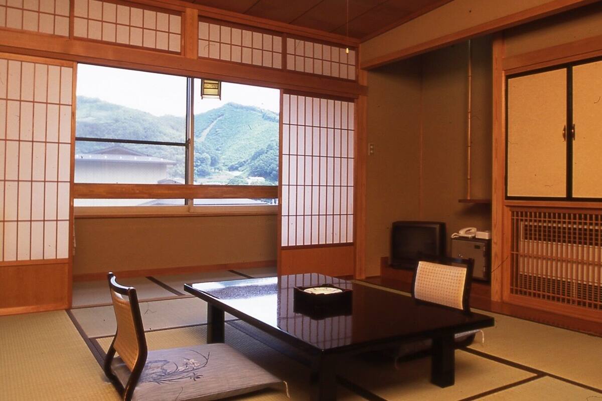 Quarto japonês tradicional com tatames, uma mesa baixa, uma cadeira, uma porta deslizante e vista para uma montanha.