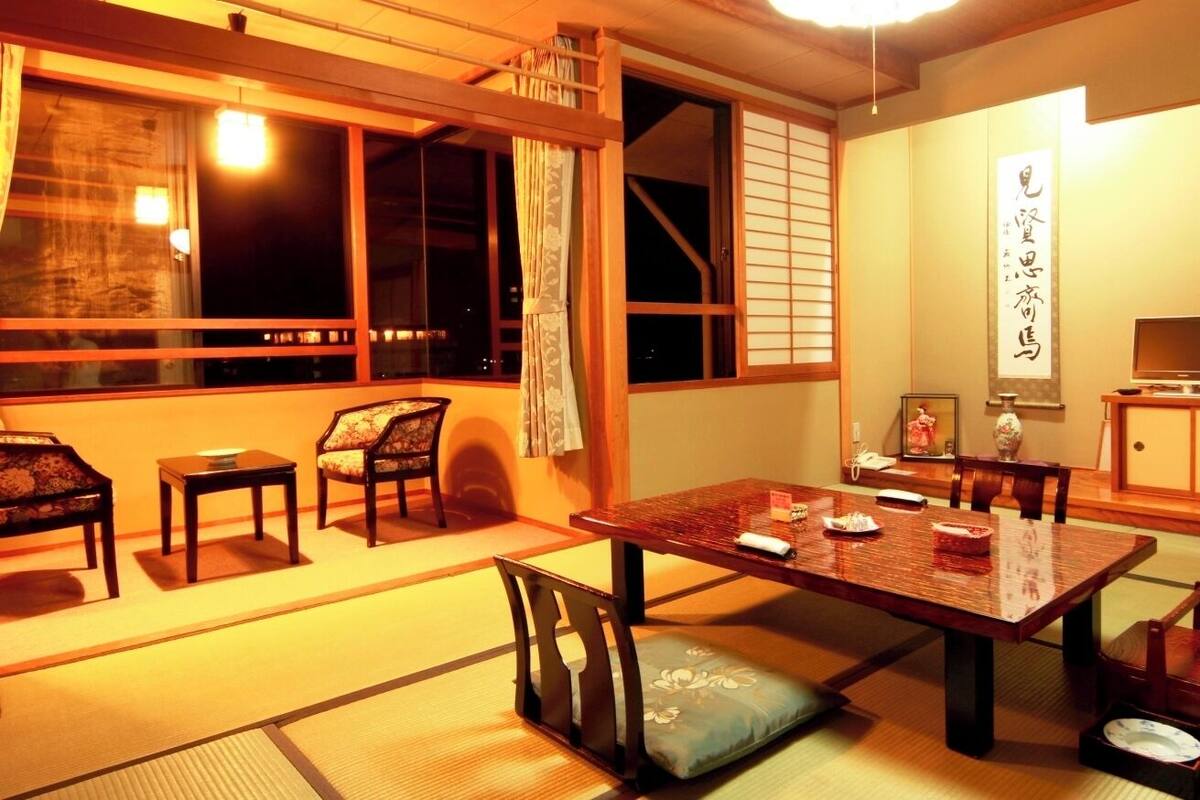 Quarto japonês tradicional com tatames, uma mesa baixa, cadeiras, televisão e uma porta deslizante.