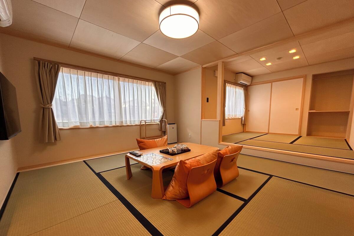Um quarto tradicional japonês com tatames, uma mesa baixa, almofadas laranja, um espelho e uma luminária no teto.