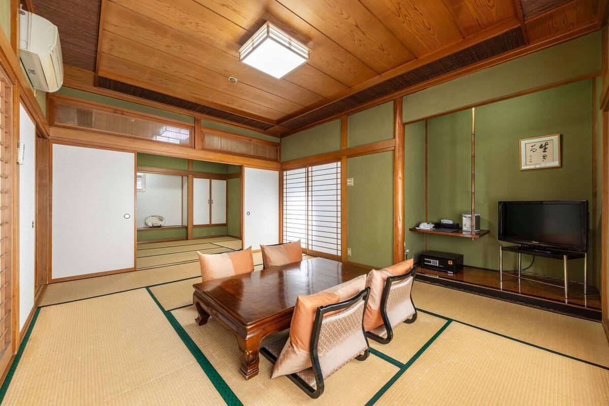 Um quarto japonês tradicional com tatames, mesa de jantar de madeira, cadeiras, uma televisão e portas corrediças.
