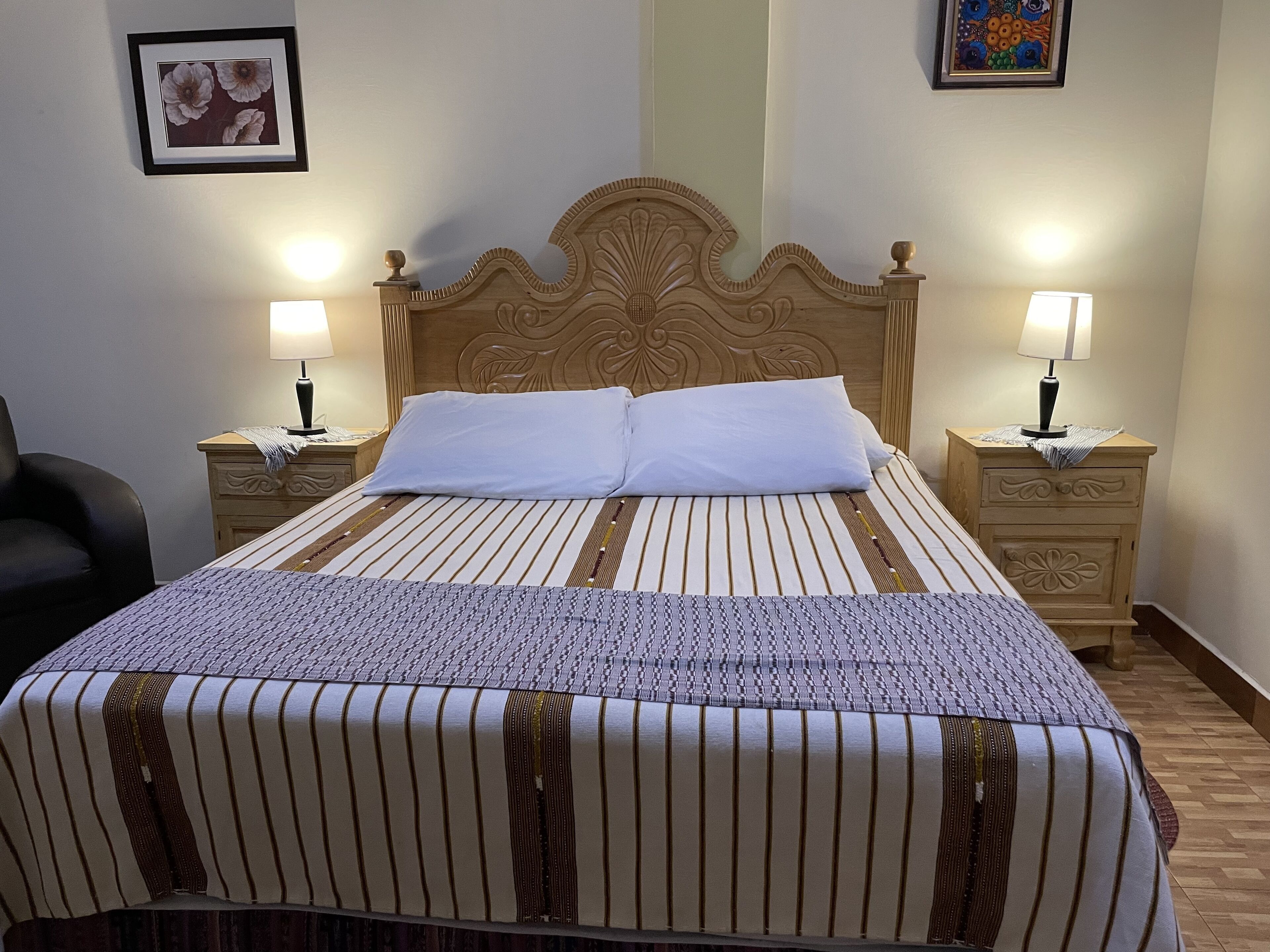 Cama bem arrumada com colcha listrada e travesseiros, ladeada por duas mesas de cabeceira com luminárias, em um quarto com cabeceira de madeira e uma moldura na parede.