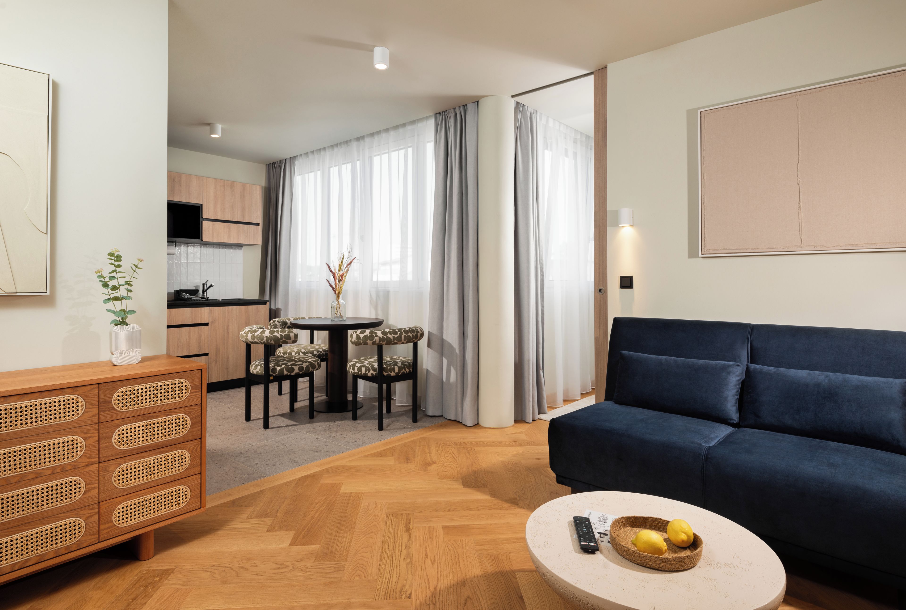Uma sala de estar moderna com um sofá azul, uma mesa de centro de madeira com frutas e um controle remoto, uma área de jantar com mesa redonda e cadeiras estampadas, e uma cozinha ao fundo.
