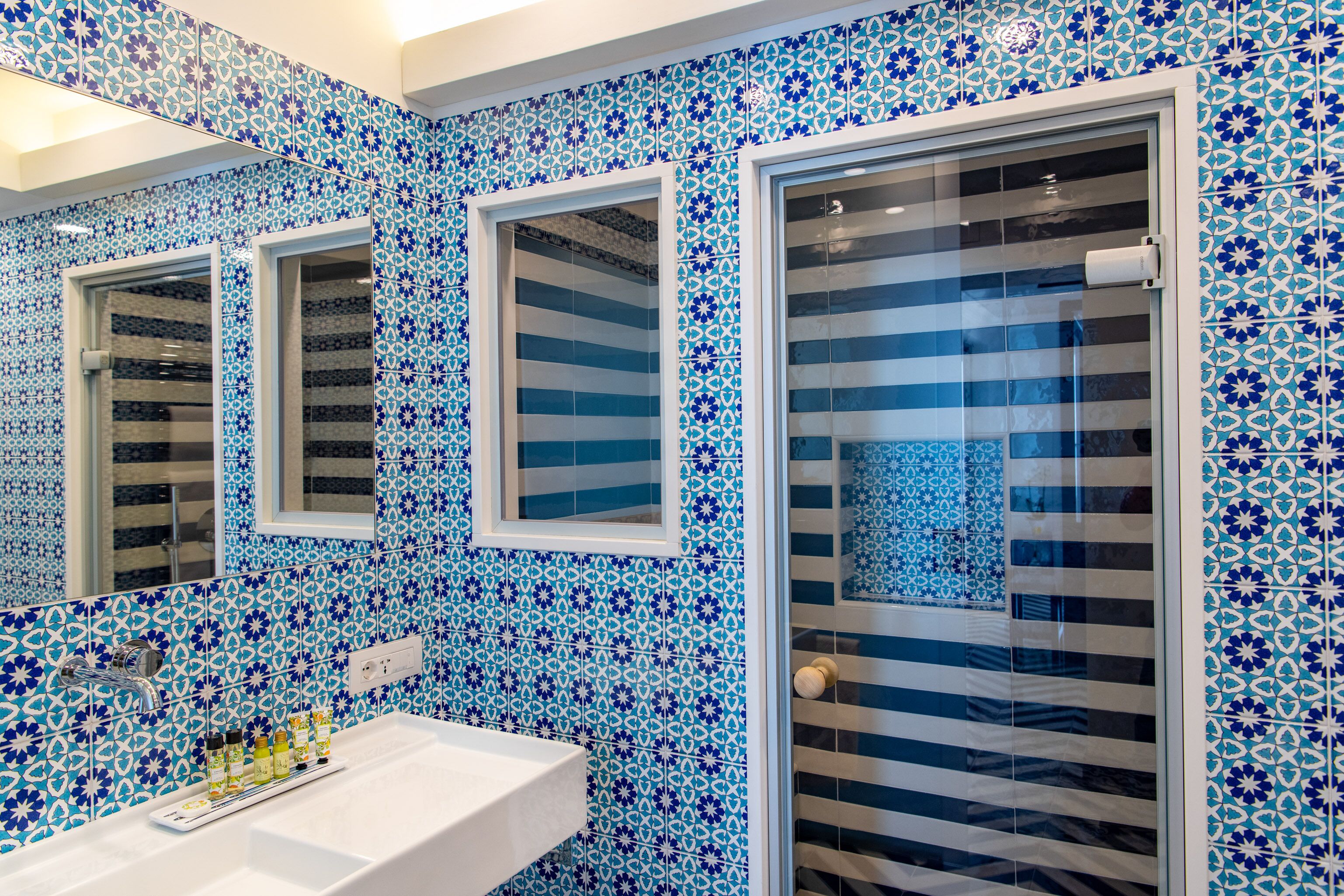 Casa de banho com azulejos padronados em azul e branco, uma pia branca e duas janelas com persianas.