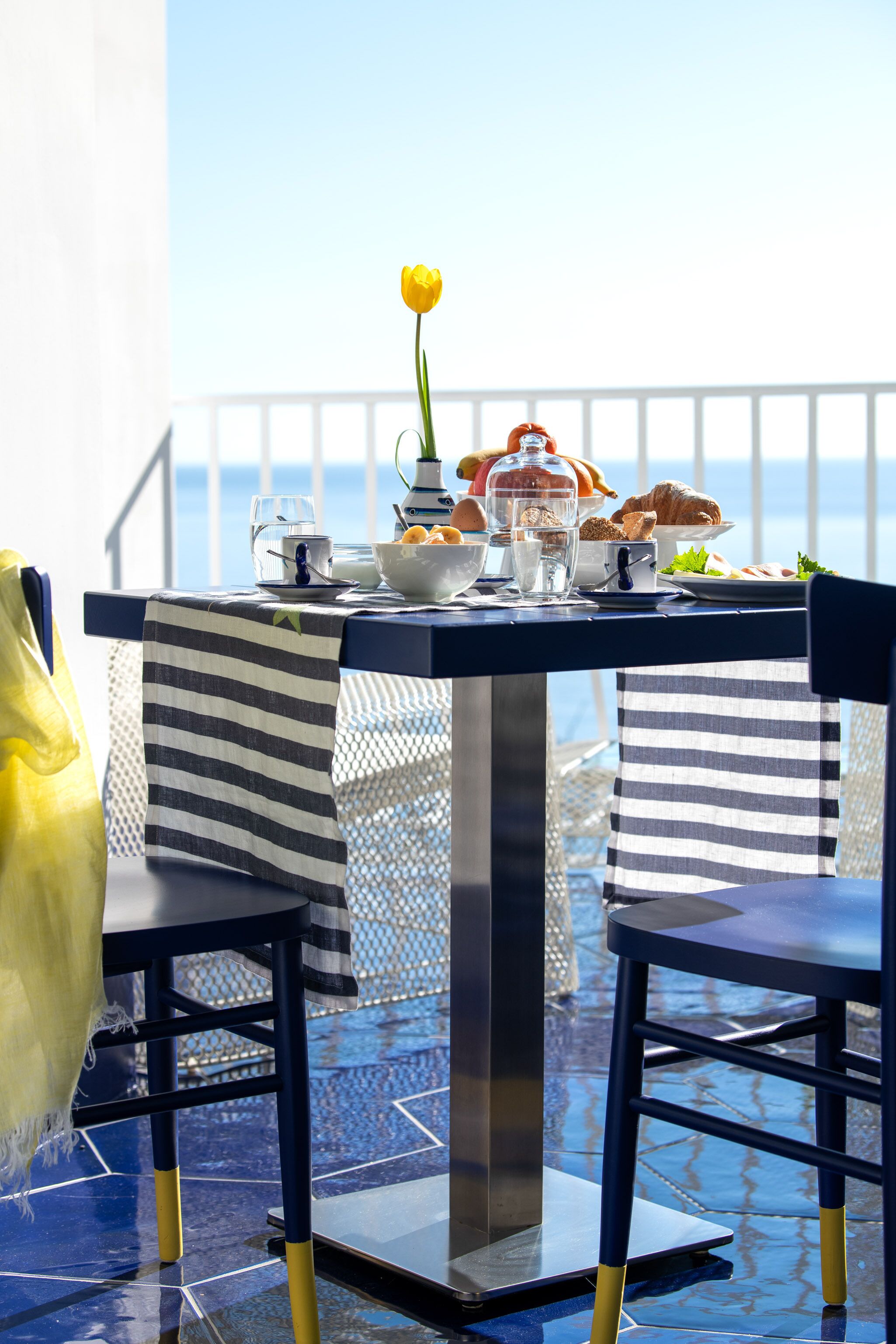 Mesa de jantar posta com itens de café da manhã, uma bolsa amarela e cadeiras listradas numa varanda com vista.