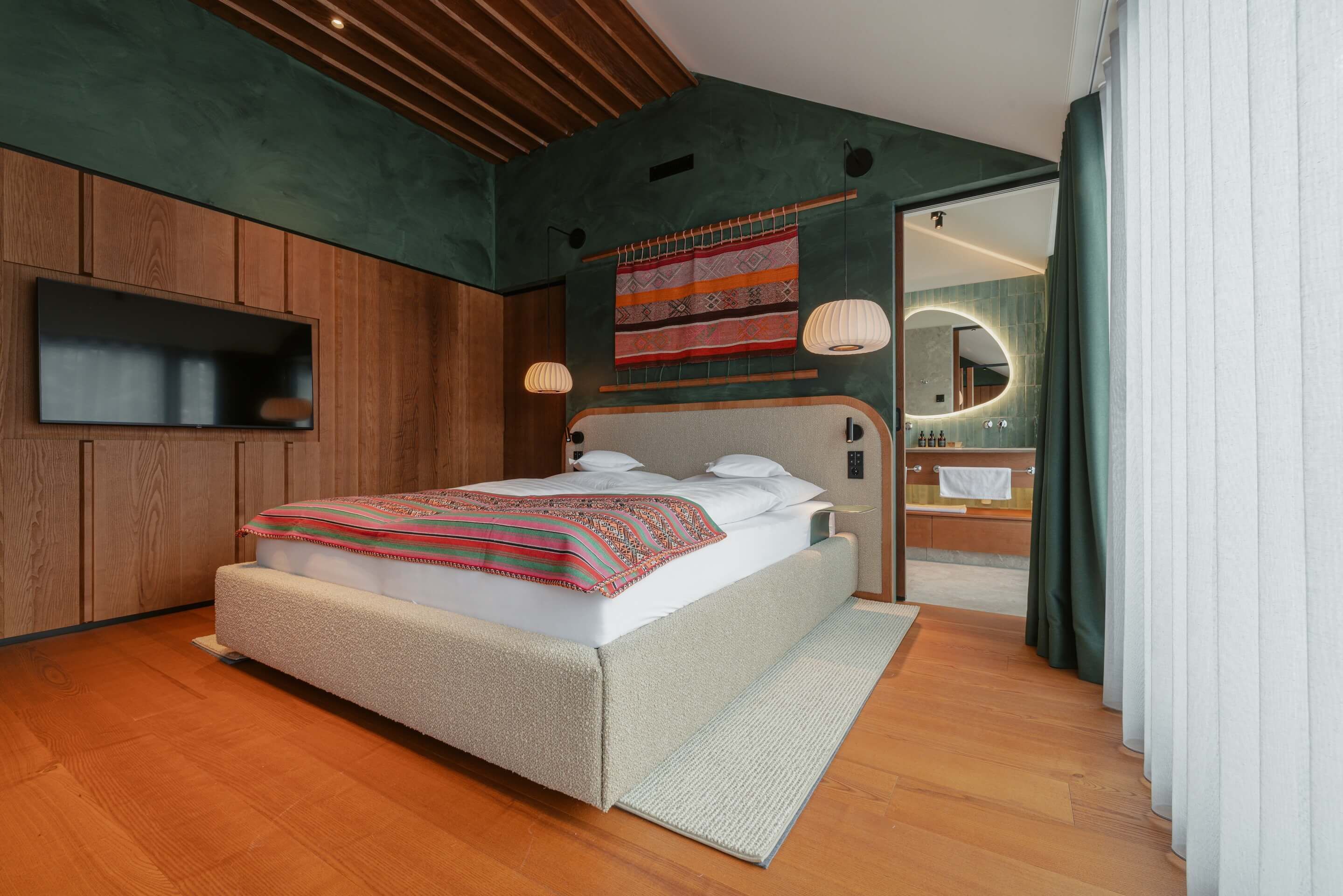 Quarto moderno com uma cama grande, painéis de madeira, TV integrada e um banheiro visível através de uma porta aberta.