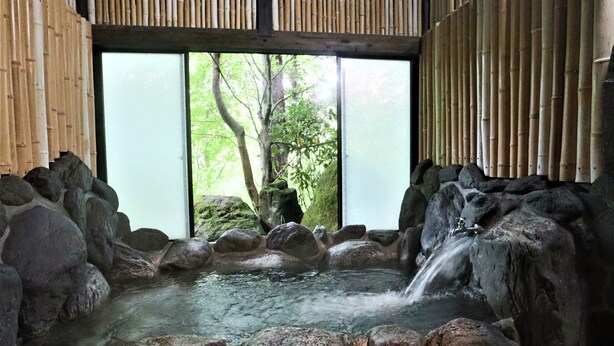 Uma área de águas termais com piscina de pedra, paredes de bambu e vista para árvores através de uma grande janela.