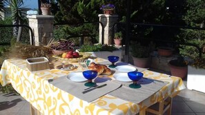 Mesa de jantar ao ar livre posta com pratos, tigelas e uma cesta, rodeada por plantas em vasos e uma parede de pedra.