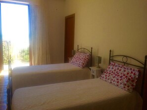 Duas camas de solteiro com roupa de cama florida, uma mesinha de cabeceira com abajur e uma janela com cortinas.