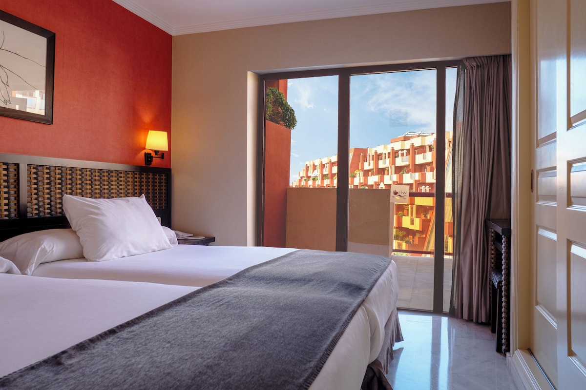 Quarto de hotel com duas camas, mesa de cabeceira, luminária, uma janela grande com vista para prédios e uma porta de vidro deslizante.