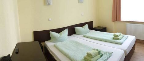 Quarto de hotel com duas camas, cada uma com um cobertor e travesseiro verdes, uma mesinha e uma cadeira.