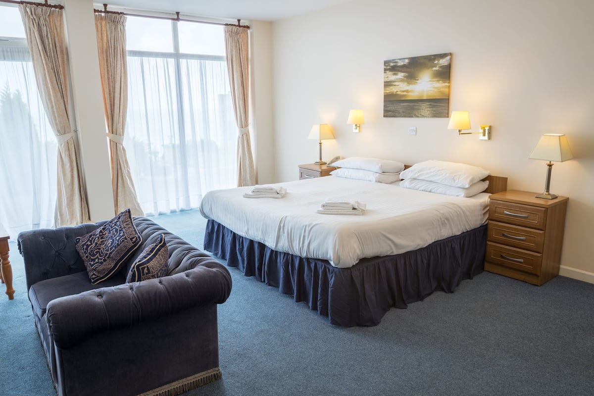Quarto de hotel com uma cama grande, duas luminárias laterais, uma mesinha de cabeceira, um aparador de madeira, um sofá de cor escura e uma janela com cortinas.