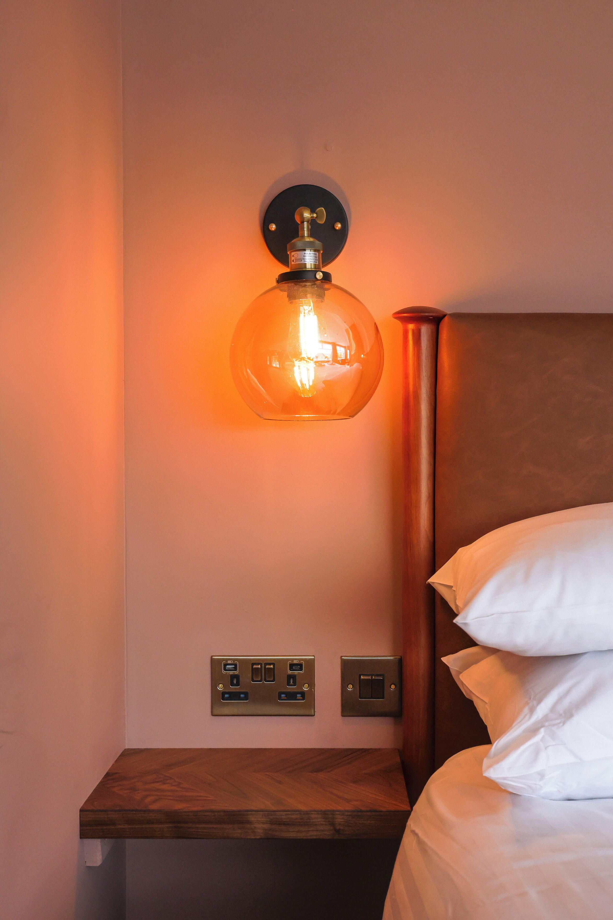 Cama bem arrumada com lençóis brancos, mesa de cabeceira de madeira com tomada e luminária de parede com cúpula de vidro transparente.