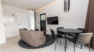 Uma sala de estar moderna com uma mesa de jantar redonda, cadeiras pretas, um sofá bege, um tapete preto e uma televisão de tela plana fixada na parede.