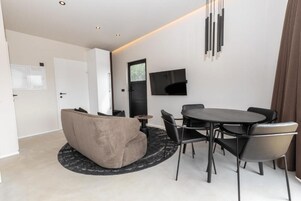 Uma sala de estar moderna com uma mesa de jantar redonda, cadeiras pretas, um sofá bege, um tapete preto e uma televisão de tela plana fixada na parede.