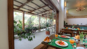 Uma área de refeições com mesas e cadeiras de madeira, um tapete verde, pratos com comida e bebidas, e vista para o jardim através de uma divisória de vidro.