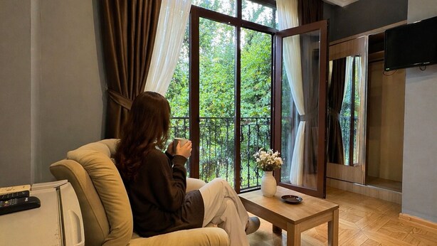Uma pessoa sentada em um sofá perto de uma janela com vista para áreas verdes, uma mesa com um vaso e uma TV fixada na parede.