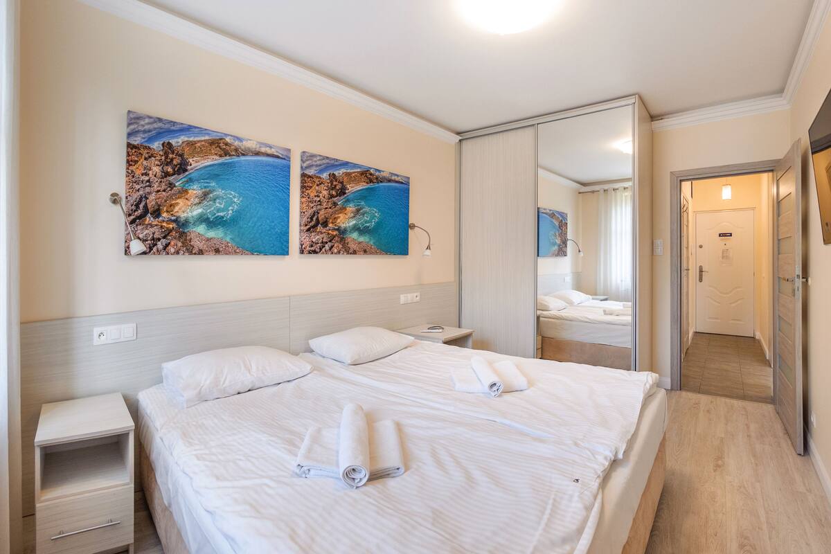 Quarto de hotel com uma cama grande, uma mesa de cabeceira e um espelho. Há duas pinturas na parede acima da cama.