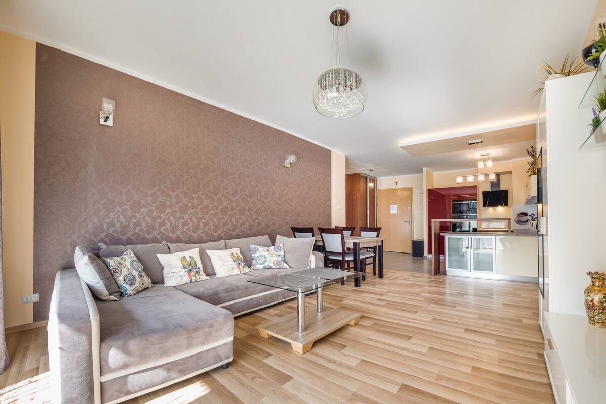 Uma sala de estar moderna com um sofá, uma mesa de centro de vidro, um candelabro e uma cozinha ao fundo.